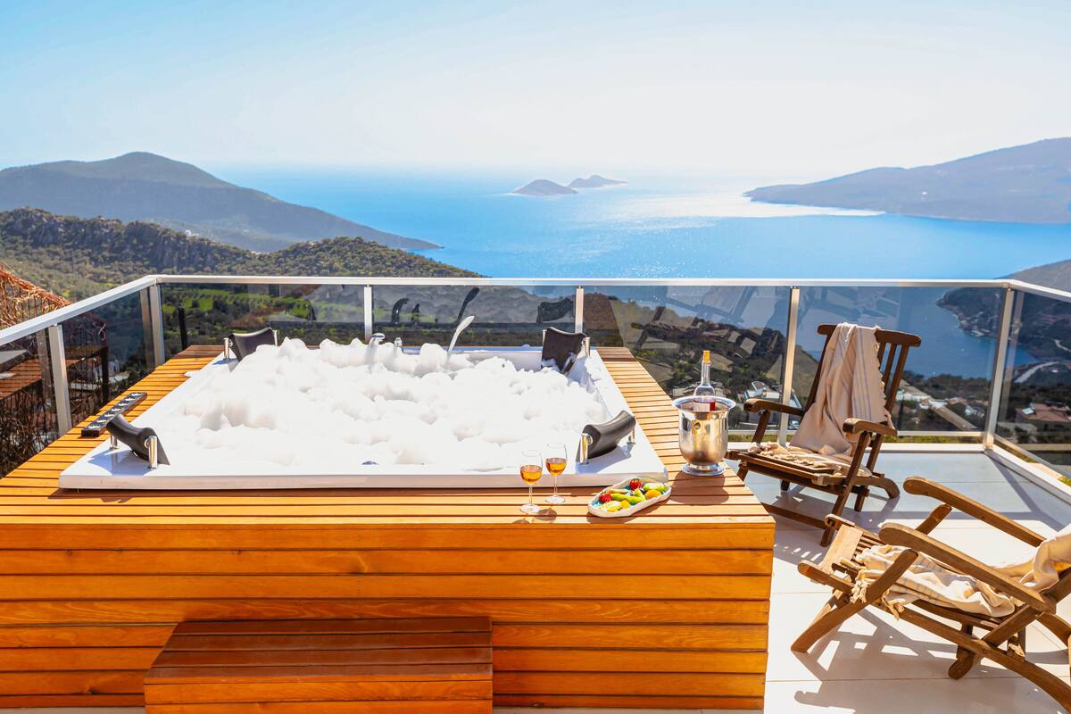 Um deck de madeira com hidromassagem, espreguiçadeiras e uma vista panorâmica do mar e colinas.
