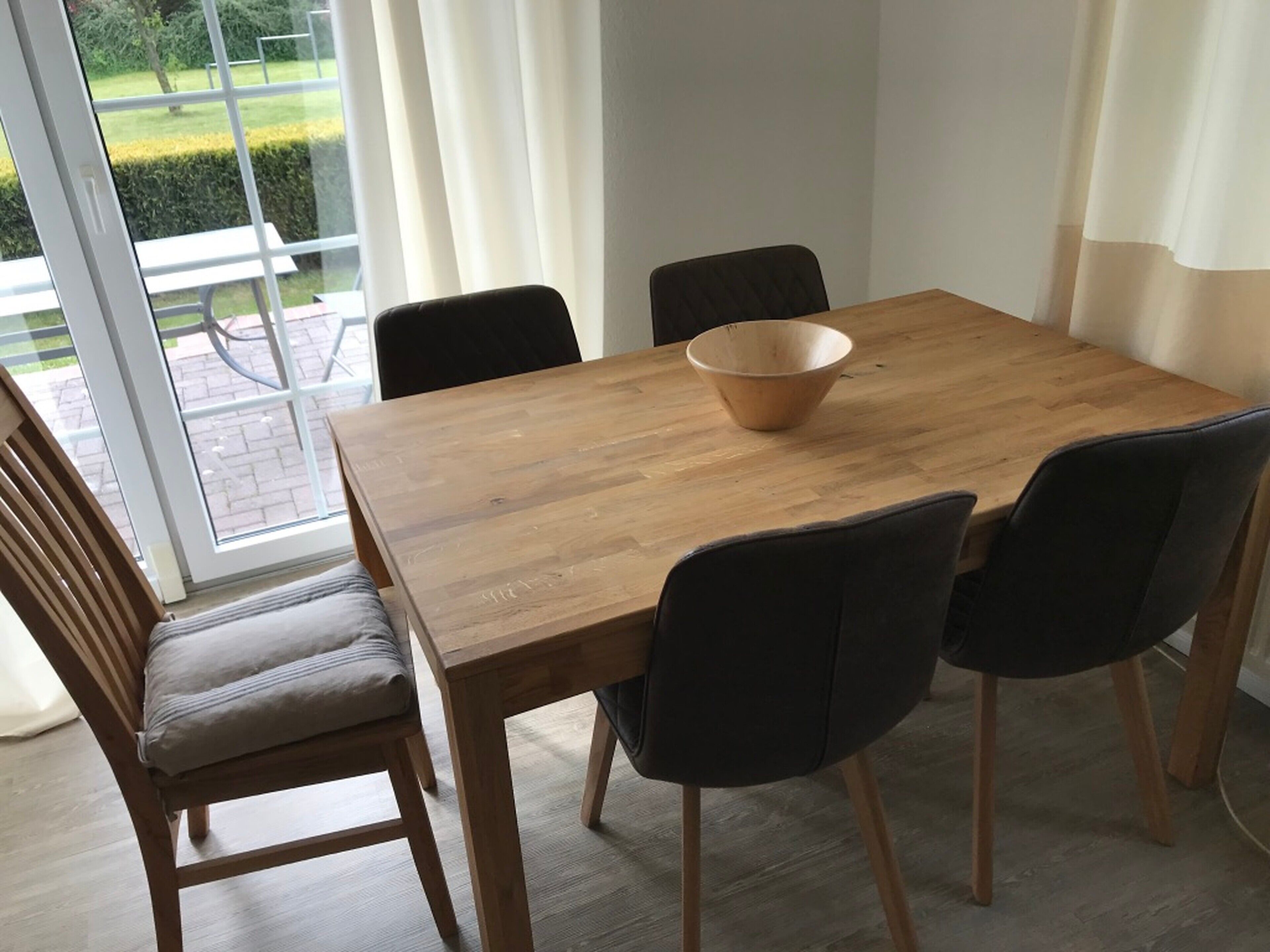 Uma mesa de jantar de madeira com uma tigela, quatro cadeiras e vista para um jardim através de uma porta de vidro.