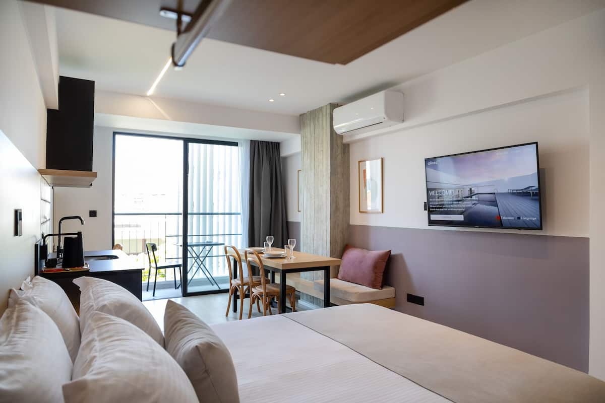 Quarto de hotel moderno com uma cama grande, área de jantar com mesa e cadeiras, televisão fixada na parede e varanda com vista.