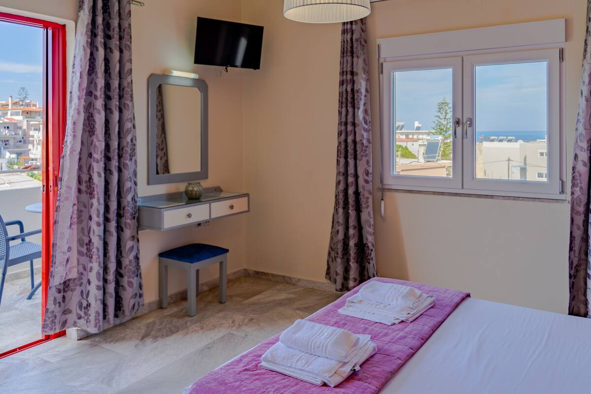Quarto de hotel com cama, escrivaninha, cadeira, televisão e janela com vista para edifícios e mar.