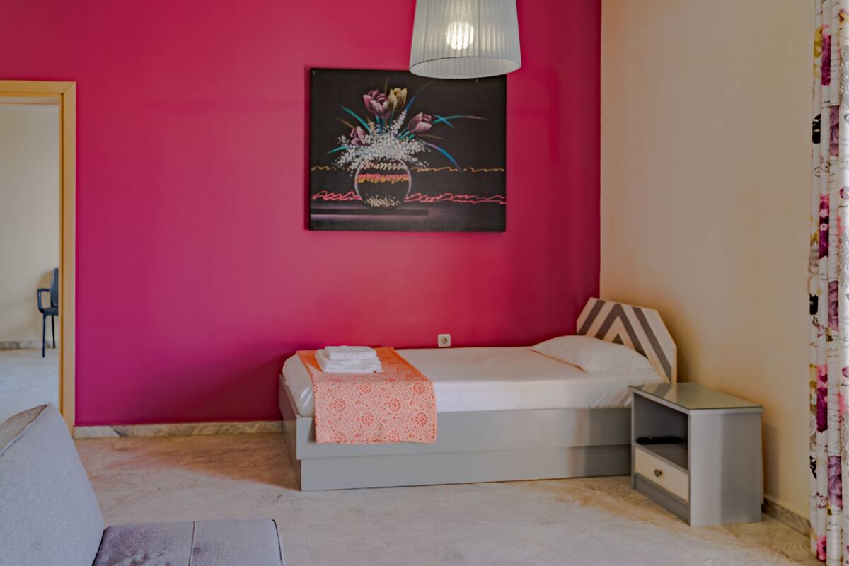 Quarto com parede rosa, cama com cabeceira branca, mesa de cabeceira e luminária fixada na parede.