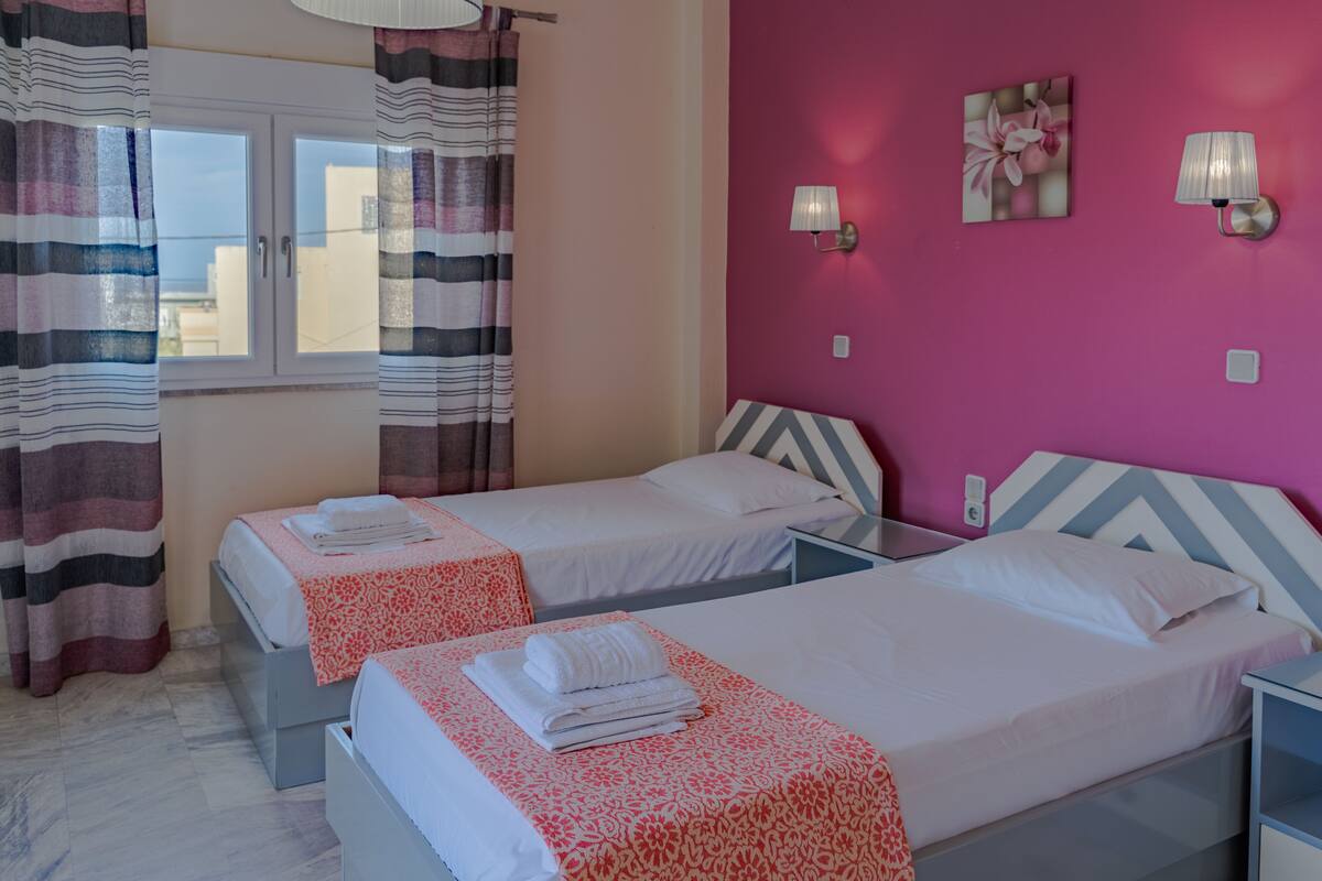 Quarto de hotel com duas camas, cortinas listradas, parede rosa e uma janela.