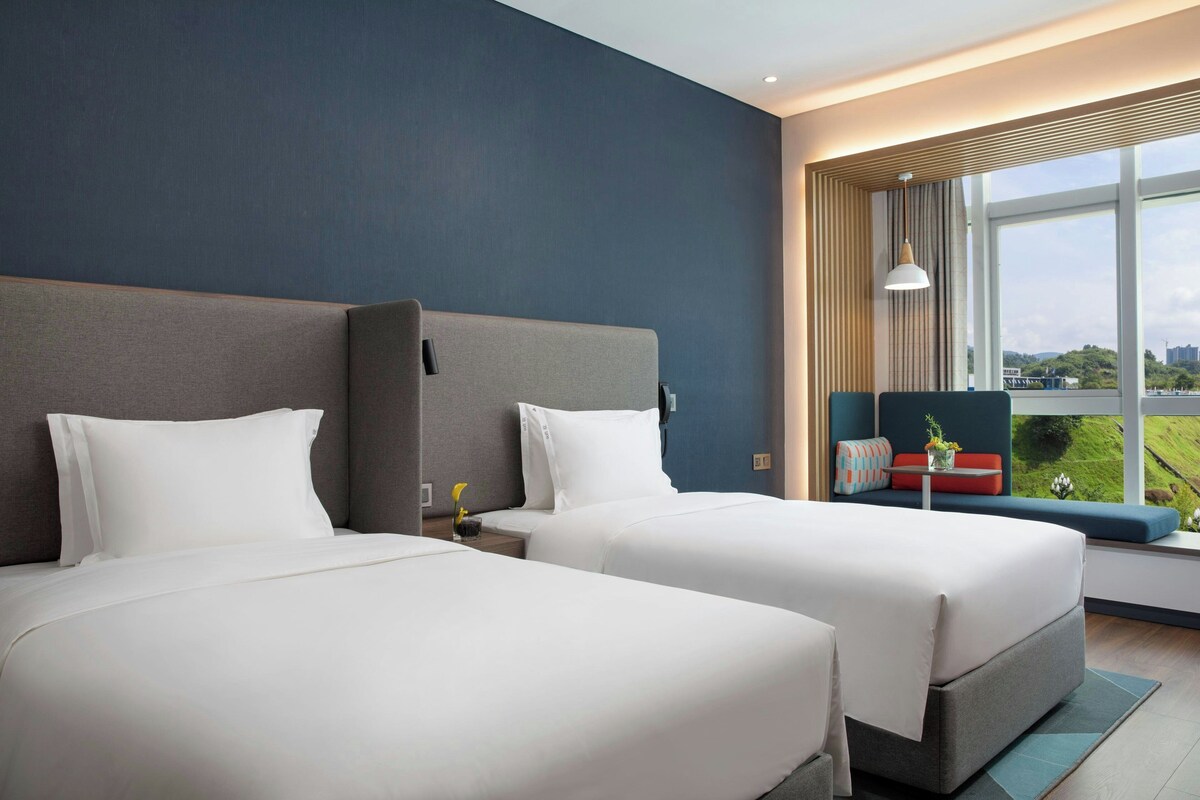 Quarto de hotel com duas camas, janela com vista para a cidade e um espaço de estar com uma planta.