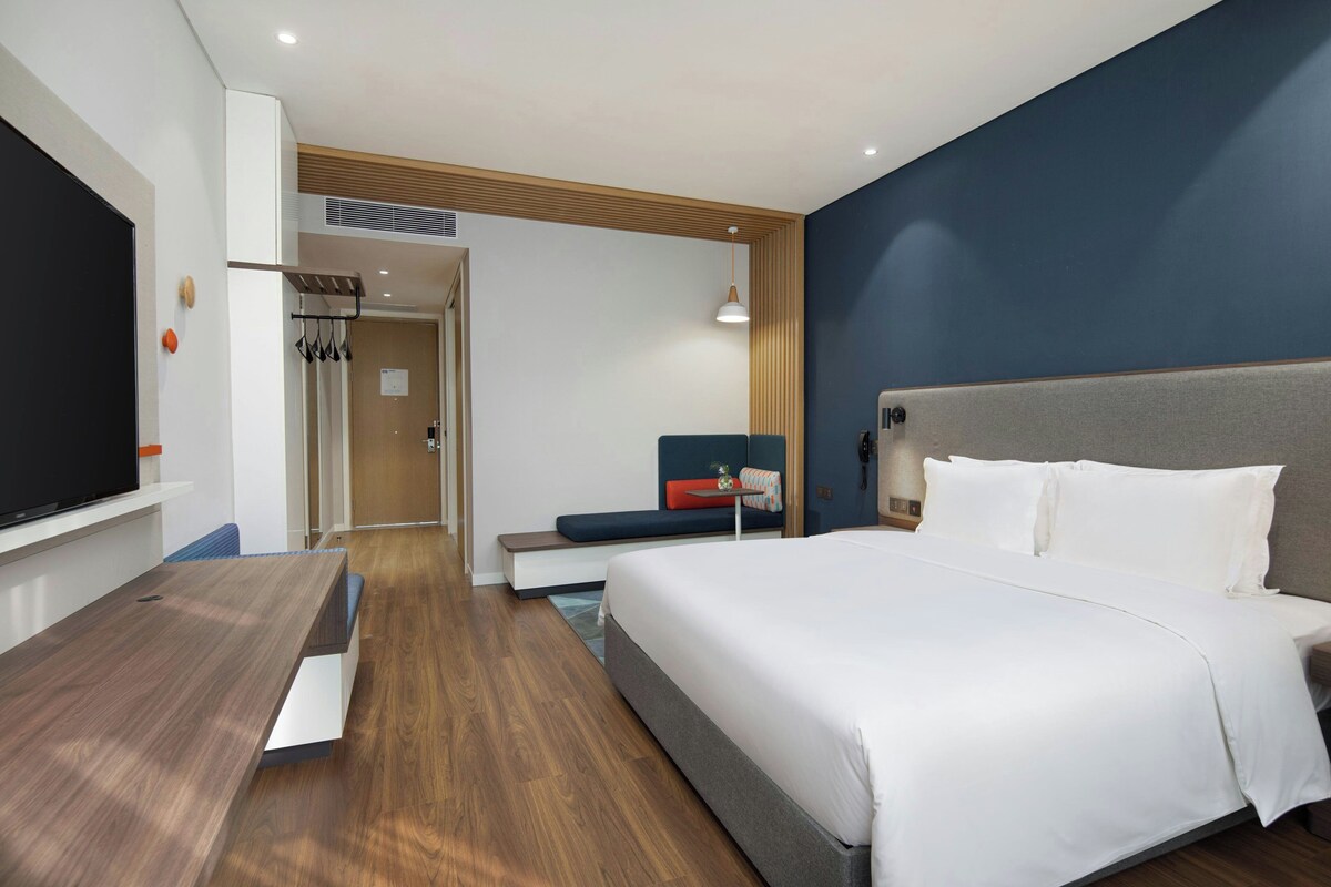 Quarto de hotel moderno com uma cama grande, televisão de tela plana, área de estar com mesa e um guarda-roupa.