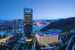 Um hotel moderno com fachada curva, situado perto de um rio e uma ponte ao fundo.