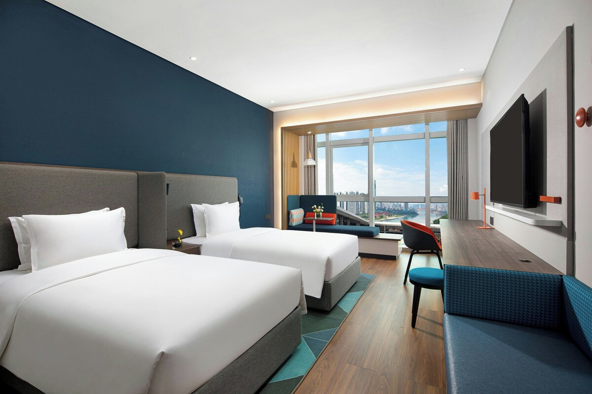 Quarto de hotel com duas camas, um sofá, uma televisão e uma grande janela com vista para a cidade.