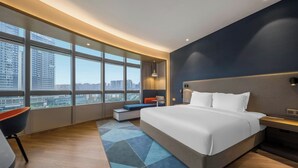 Quarto de hotel moderno com uma cama grande, área de estar, escrivaninha e vista da cidade através de amplas janelas.