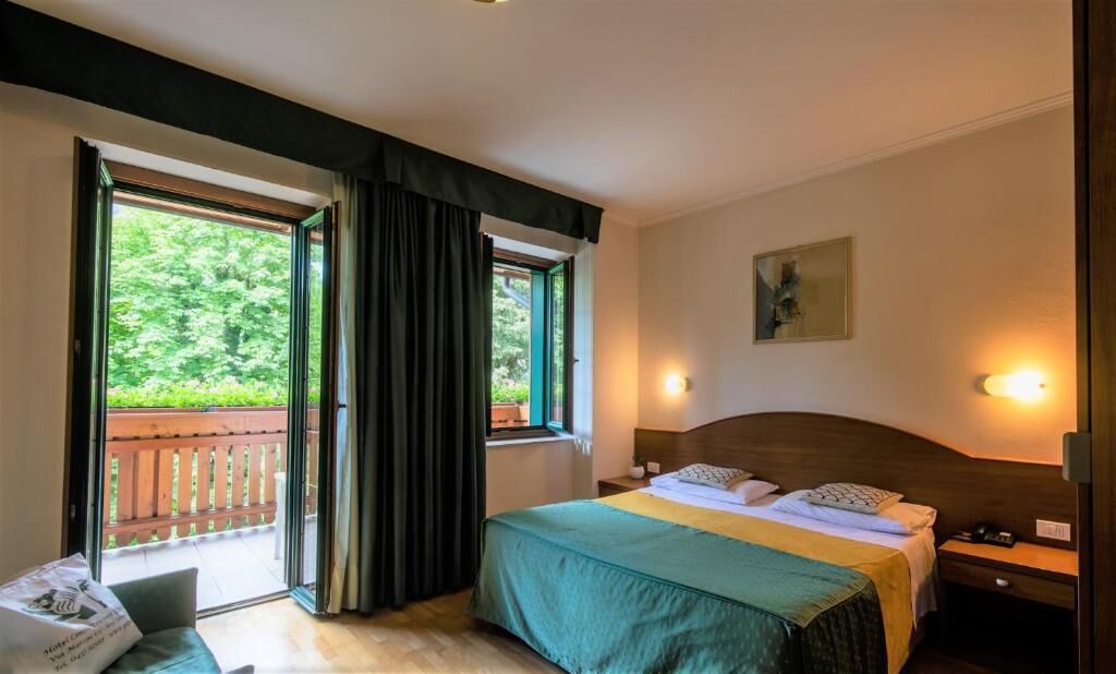 Quarto com cama, duas mesas de cabeceira, um criado-mudo, janela com cortinas e vista para o verde lá fora.