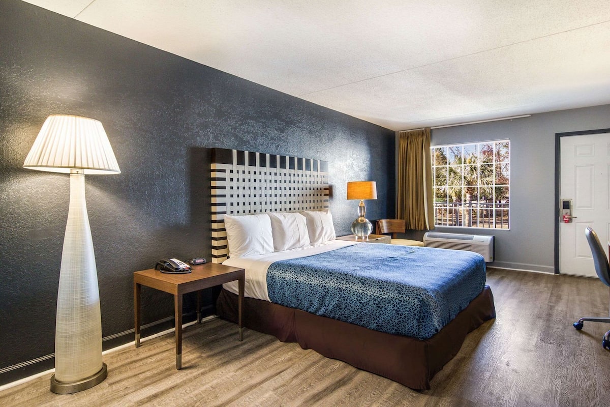Quarto de hotel com cama, mesa de cabeceira, abajur e janela com vista.