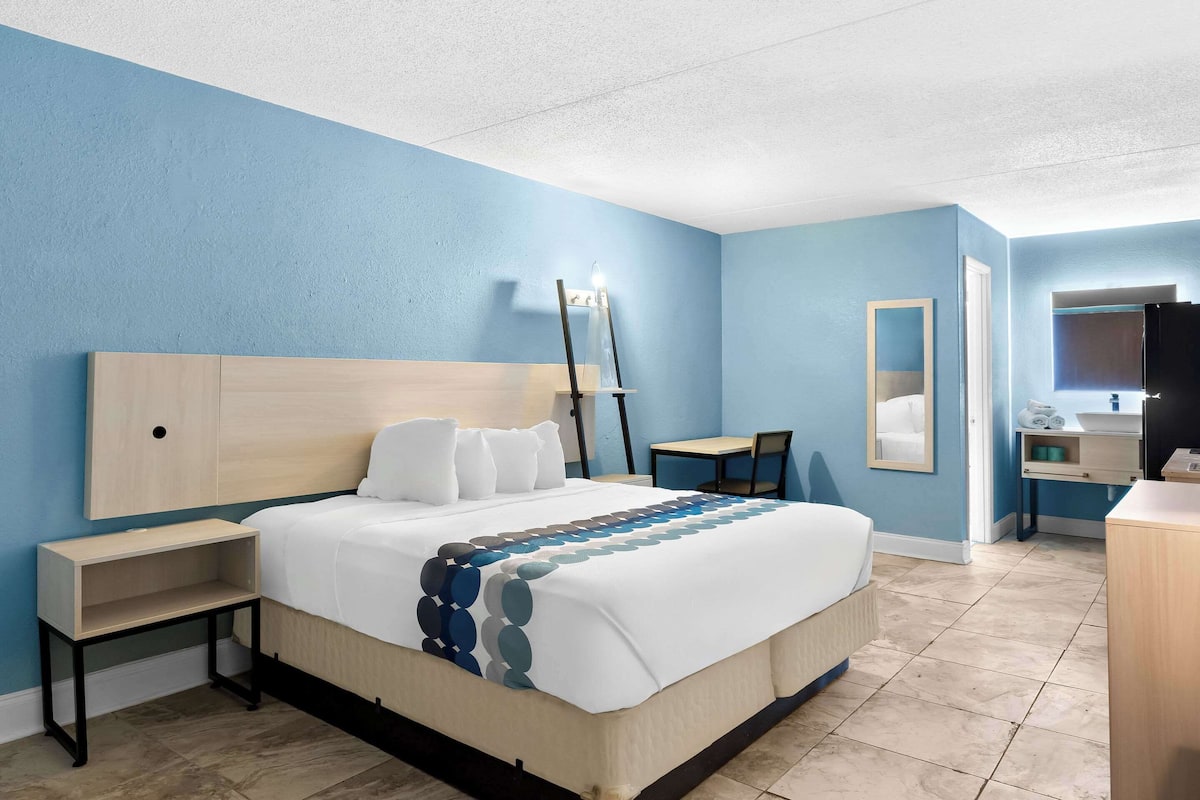 Quarto de hotel com cama, mesas de cabeceira, escrivaninha, cadeira, espelho e televisão.