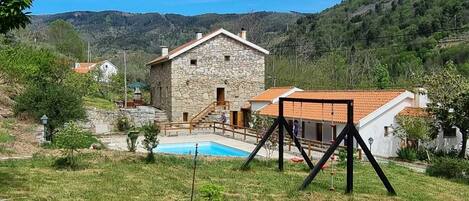 Um edifício de pedra com piscina e baloiço numa área gramada, com colinas ao fundo.