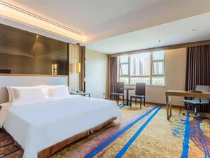 Quarto de hotel com uma cama grande, uma escrivaninha e uma cadeira. Há uma janela com vista para edifícios.