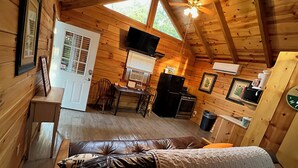 Uma cabana acolhedora com interior de madeira, uma cama, uma escrivaninha, uma televisão e uma pequena cozinha.