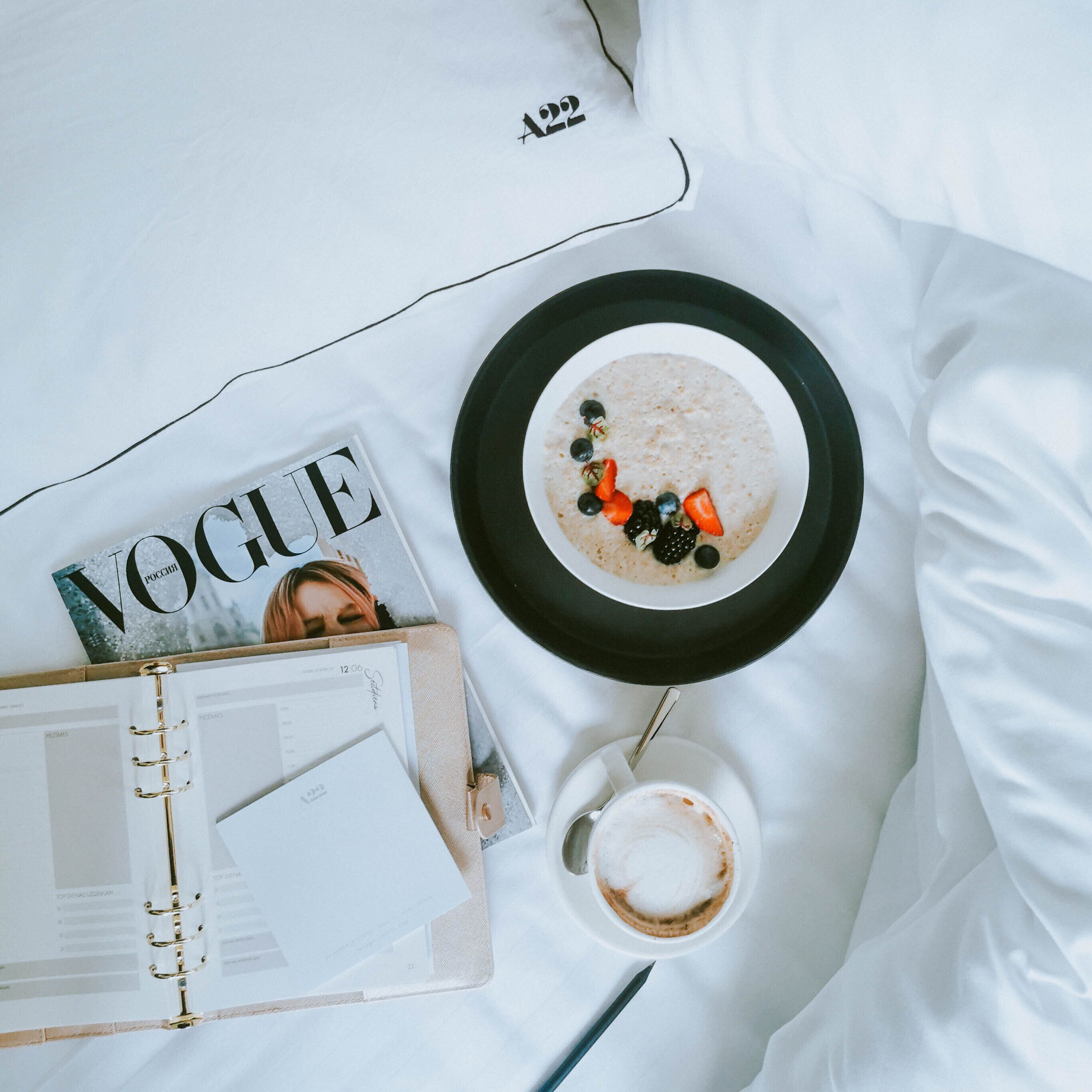 Cama bem arrumada com uma revista Vogue, uma tigela de aveia com frutas vermelhas e uma xícara de café.