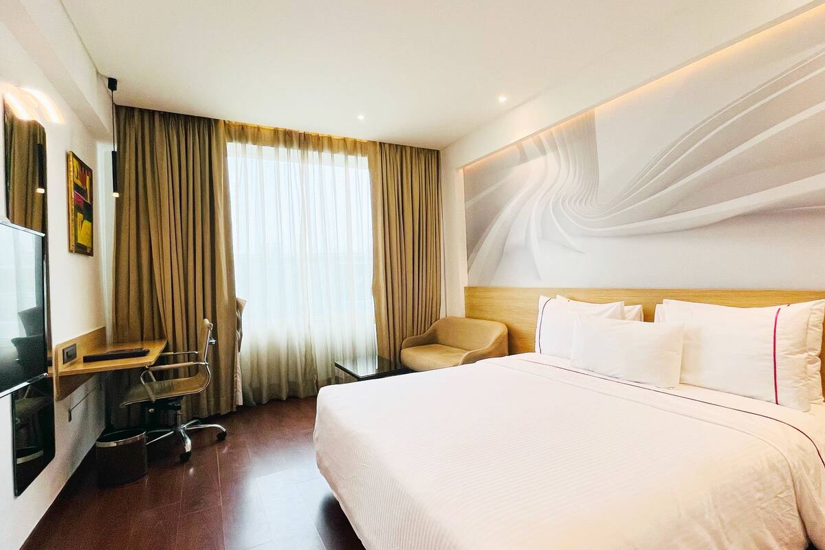 Quarto de hotel com cama, escrivaninha, cadeira, televisão e telefone.