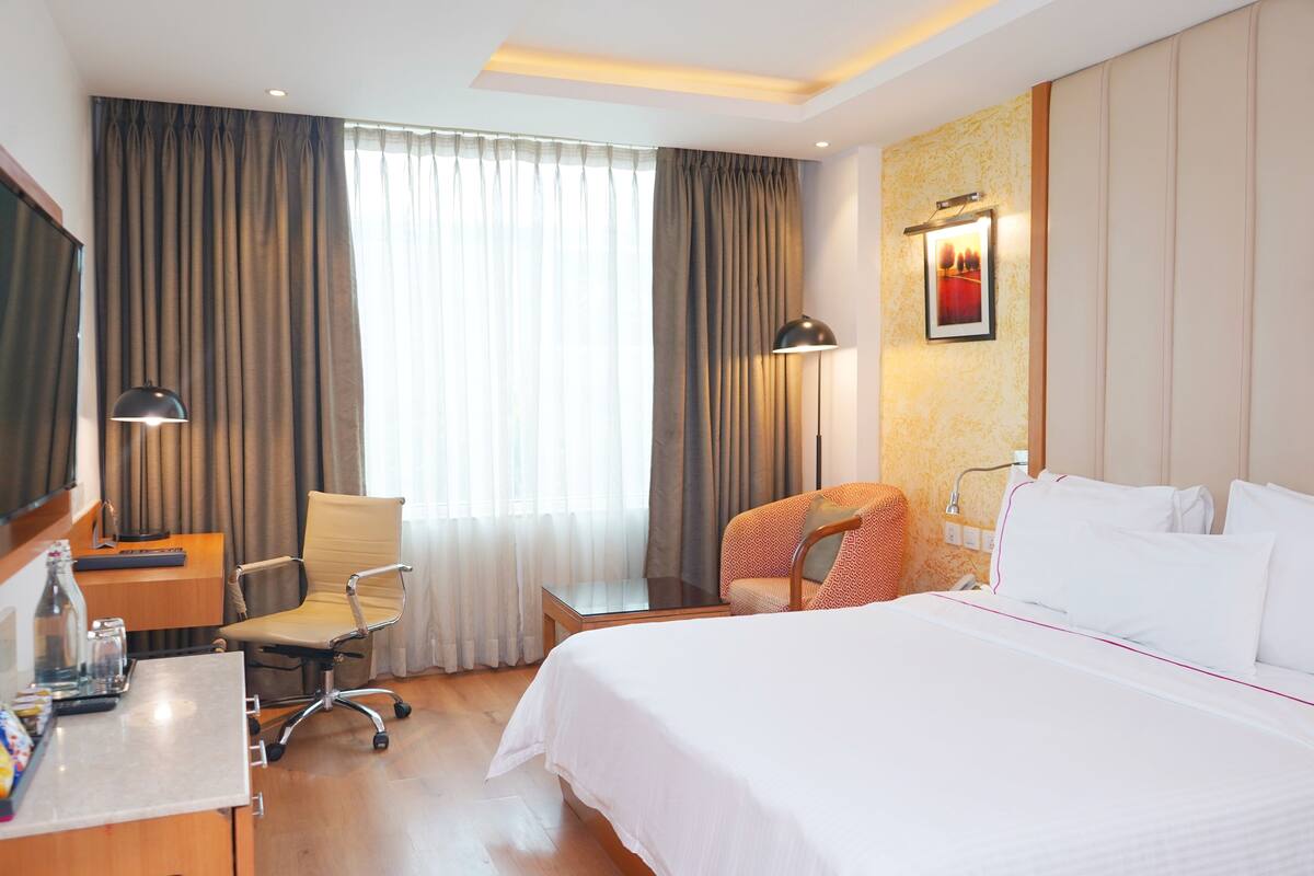Quarto de hotel com uma cama grande, uma escrivaninha com cadeira, uma mesinha e um abajur.