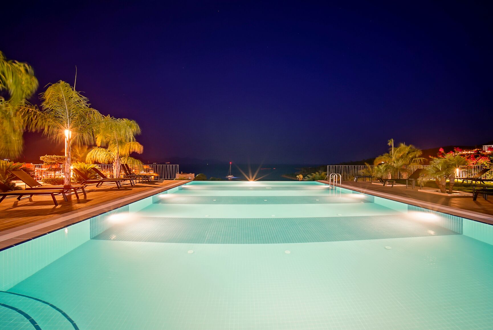 Piscina à noite com espreguiçadeiras e palmeiras, cercada por uma grade e vista para o oceano.