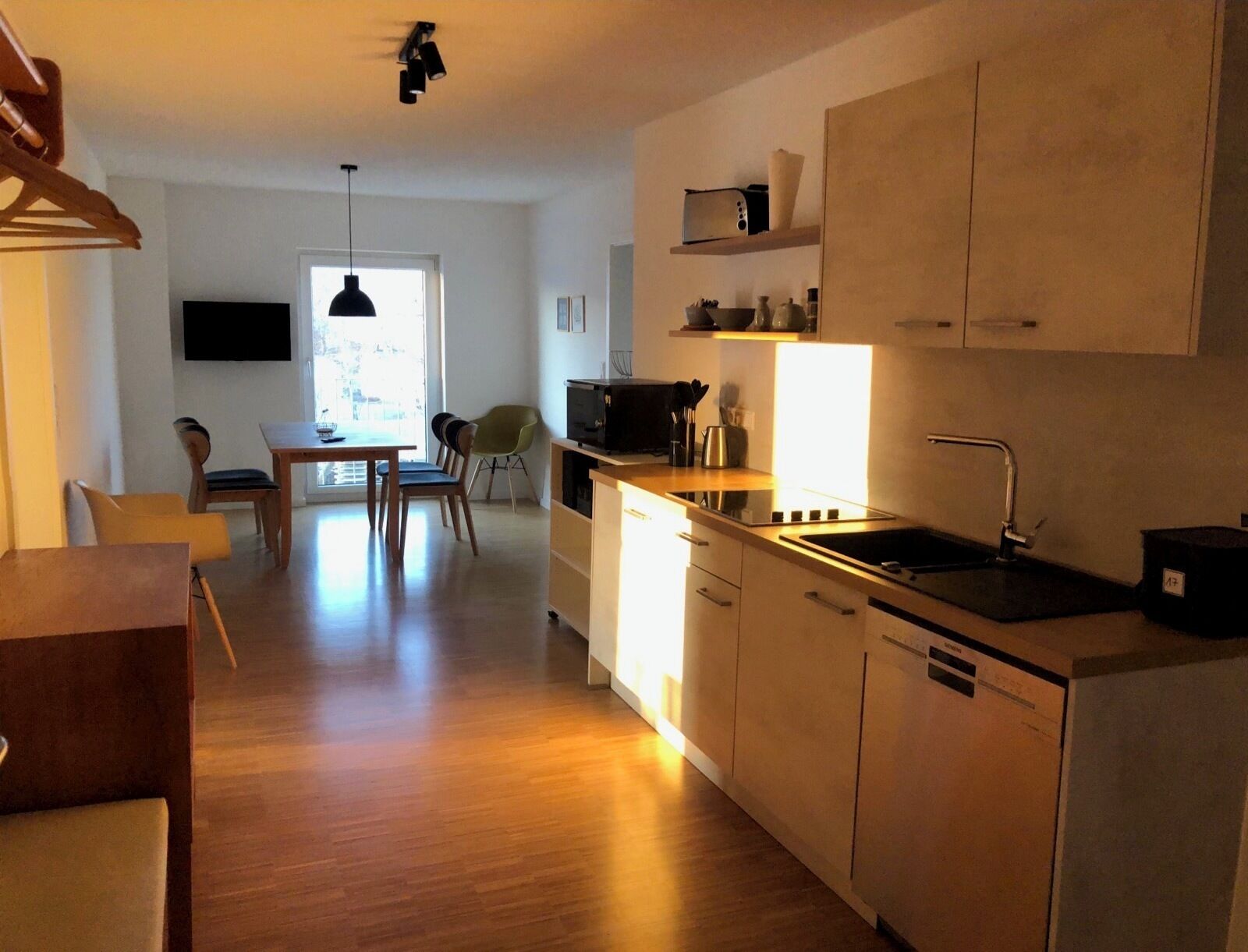 Cozinha moderna com armários de madeira, micro-ondas embutido e pia. Há uma área de jantar com mesa e cadeiras, e uma grande janela que proporciona iluminação natural.