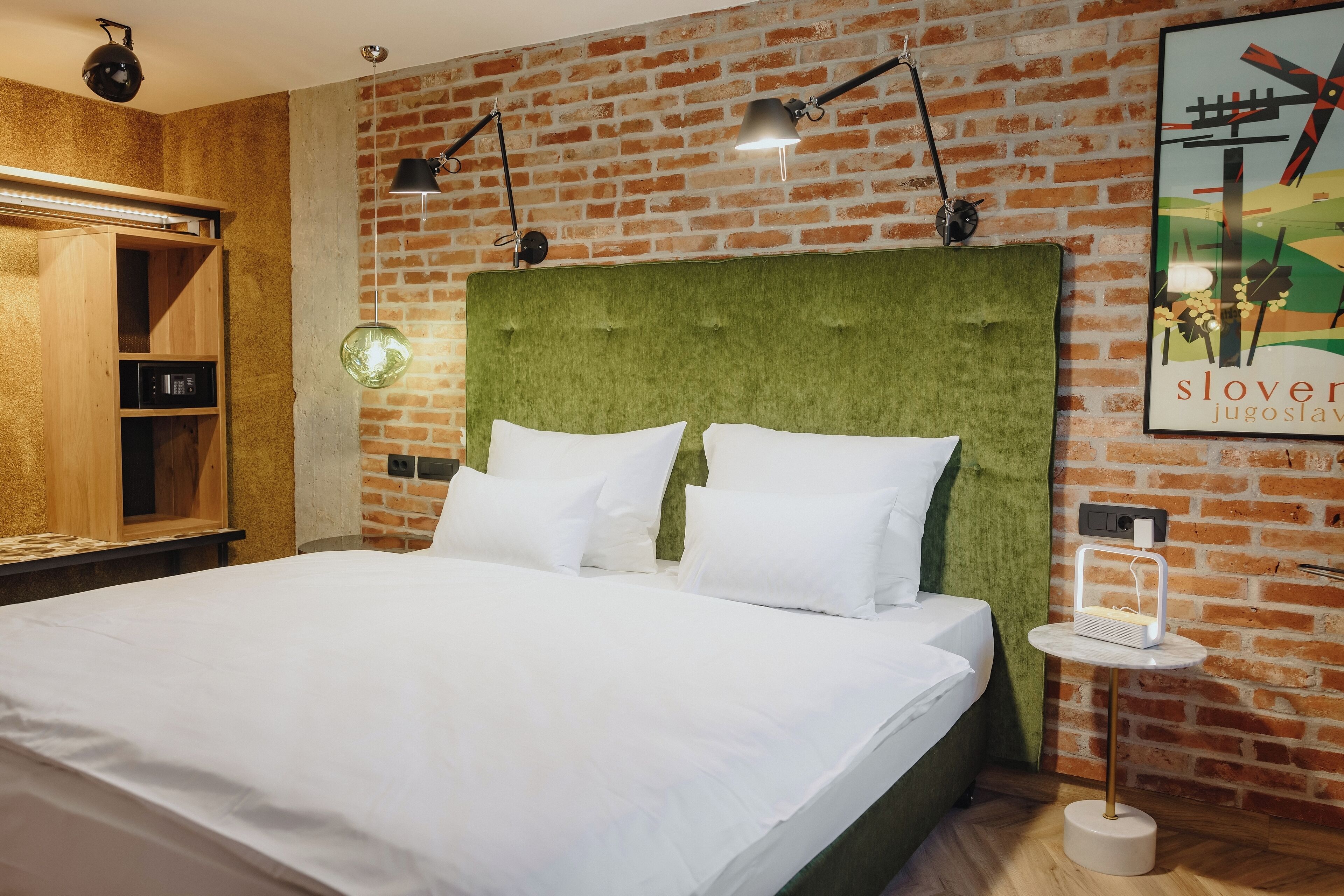 Quarto de hotel com parede de tijolos, cabeceira estofada verde, roupa de cama branca, mesa de cabeceira com abajur e um quadro na parede.