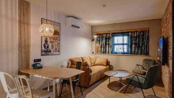 Uma sala de estar moderna com um sofá, uma mesa de jantar e duas cadeiras. Há uma janela com cortinas, um lustre pendente e um ar condicionado fixado na parede.
