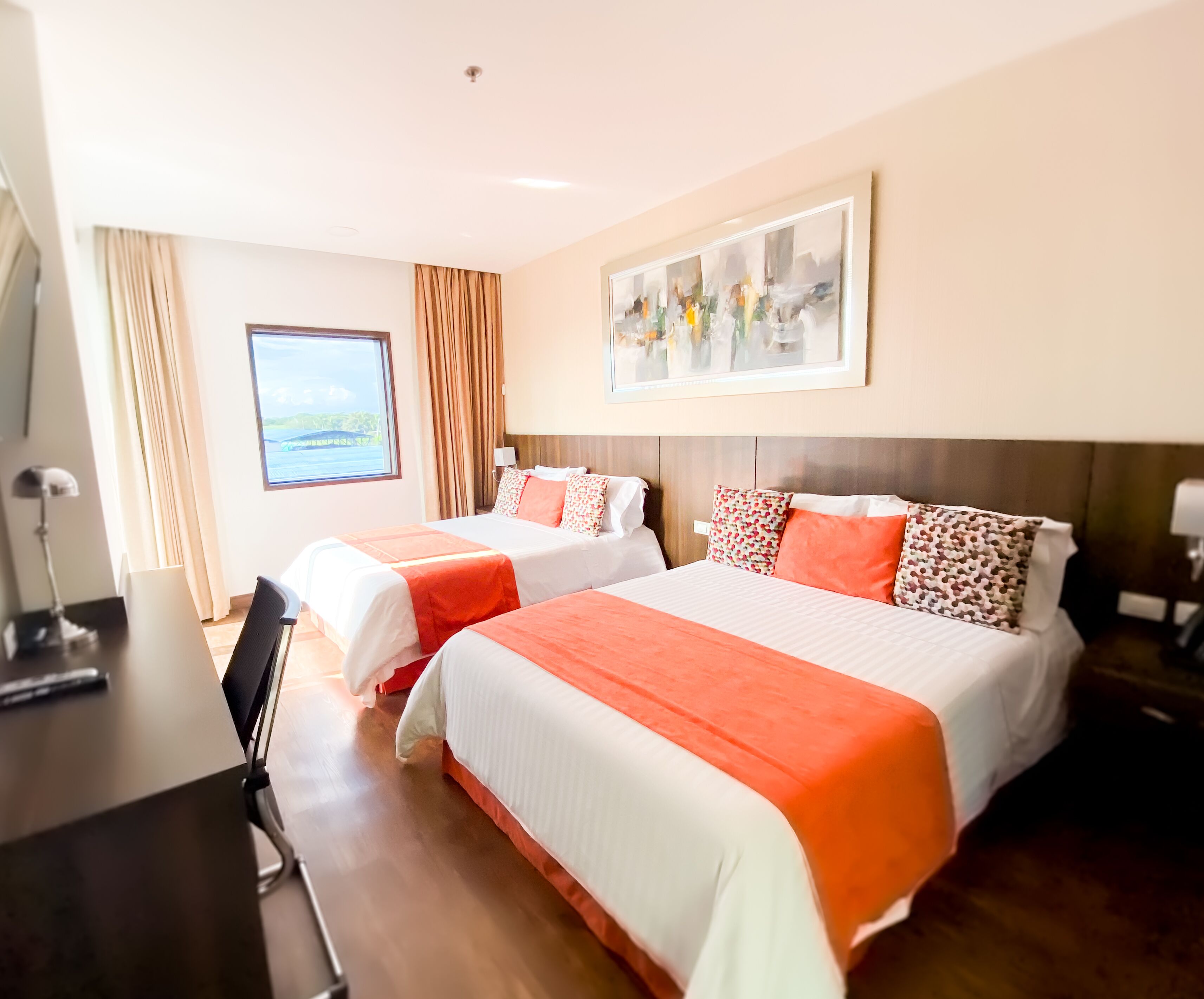 Quarto de hotel com duas camas, uma escrivaninha, uma cadeira, janela com vista e um quadro na parede.
