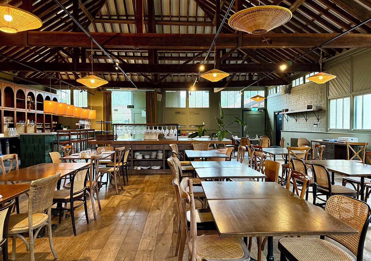 Um restaurante amplo com mesas e cadeiras de madeira, janelas grandes e luminárias de vime penduradas.