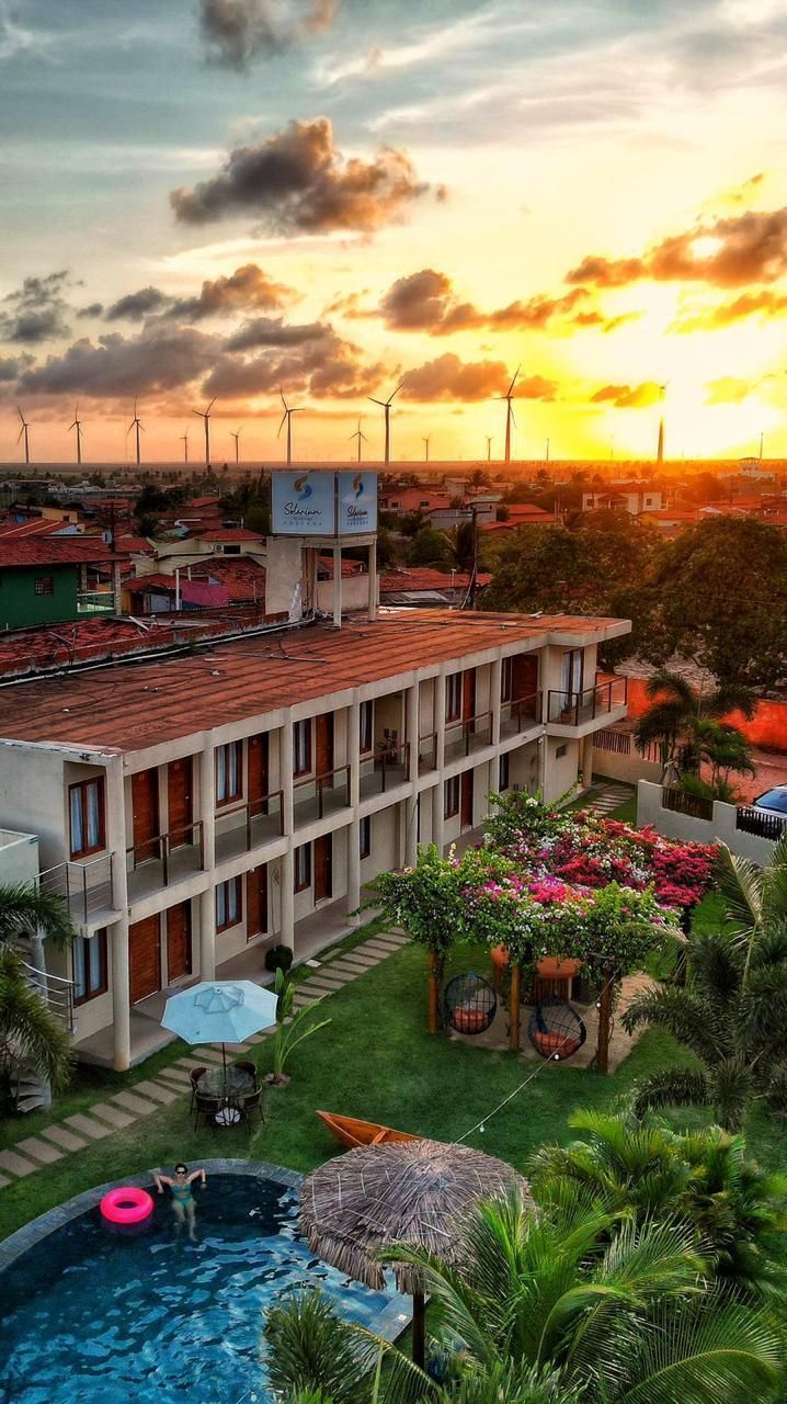 Hotel com piscina no terraço, rodeado por vegetação exuberante e vista para turbinas eólicas ao longe.