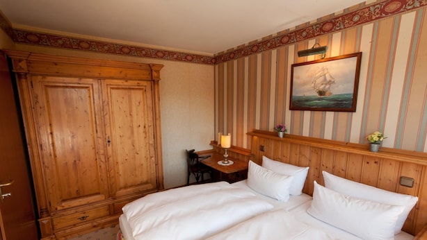 Quarto de hotel com um armário de madeira grande, uma cama com roupa de cama branca, uma escrivaninha com luminária e um quadro de um navio a vela na parede.