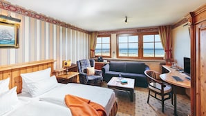 Quarto de hotel com cama, sofá, escrivaninha e vista para o mar através de grandes janelas.