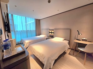 Quarto de hotel com duas camas, uma mesa de trabalho com cadeira, televisão e mesa de cabeceira com garrafa de água e caixa de lenços.