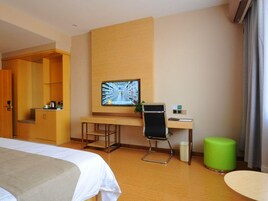 Quarto de hotel com uma cama, uma escrivaninha, uma cadeira, uma mesinha, um cesto de lixo e um banquinho verde.