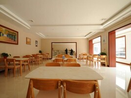 Uma sala de jantar ampla, com mesas e cadeiras de madeira, uma cozinha ao fundo e grandes janelas que permitem a entrada de luz natural.