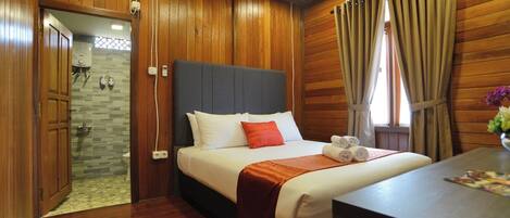 Quarto de hotel com painéis de madeira, cama com roupa de cama branca e vermelha, mesa de cabeceira com toalhas e janela com cortinas.