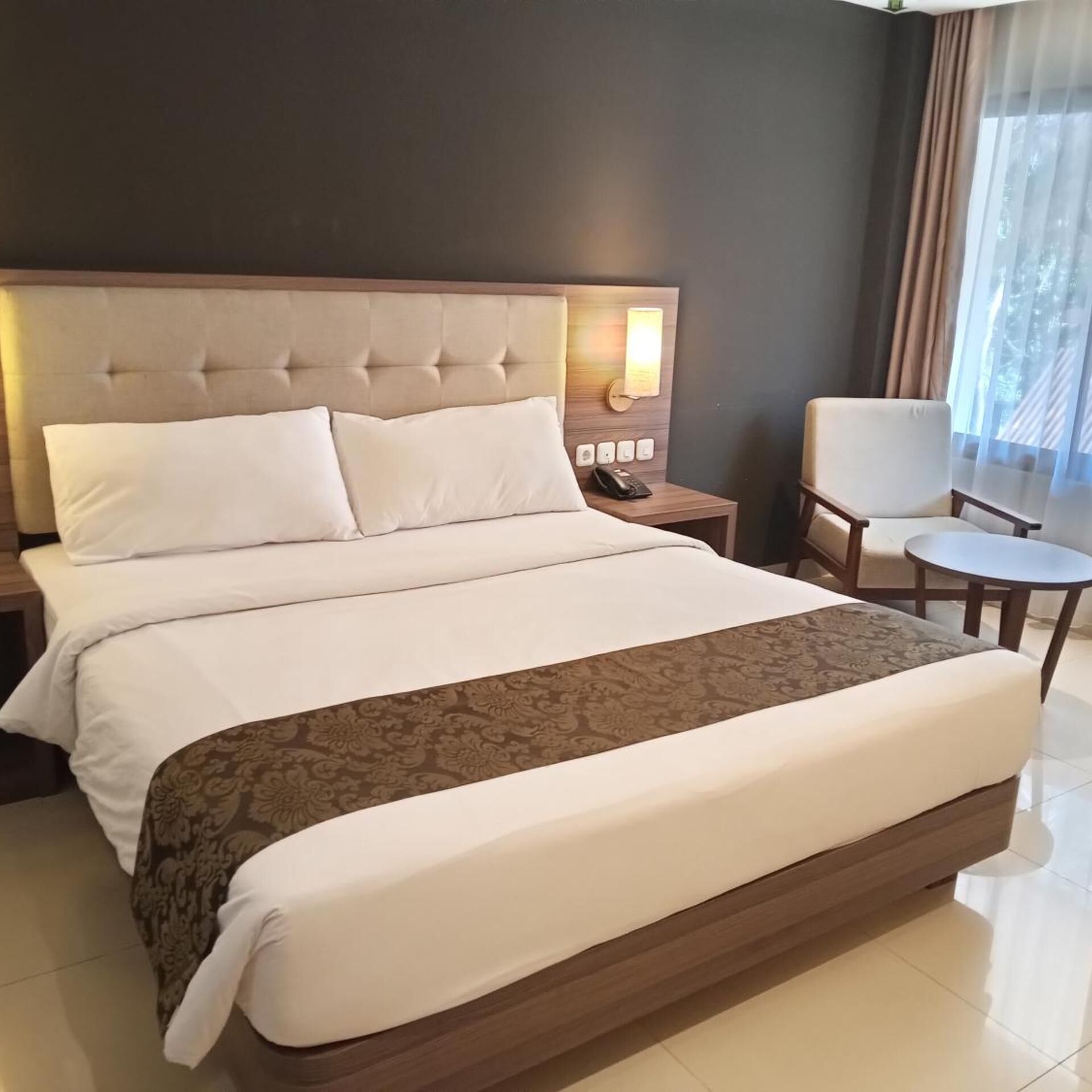 Quarto de hotel com uma cama grande, uma mesa de cabeceira com abajur, uma cadeira e vista para o exterior.