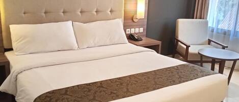 Quarto de hotel com uma cama grande, uma mesa de cabeceira com abajur, uma cadeira e vista para o exterior.