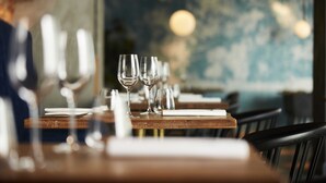 Interior de restaurante com mesas preparadas para refeições, com copos e pratos transparentes.