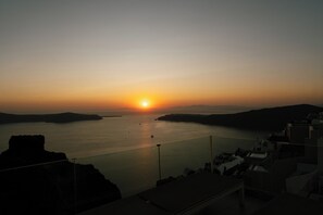 Vista do pôr do sol de uma varanda com vista para um mar calmo e formações terrestres ao longe.