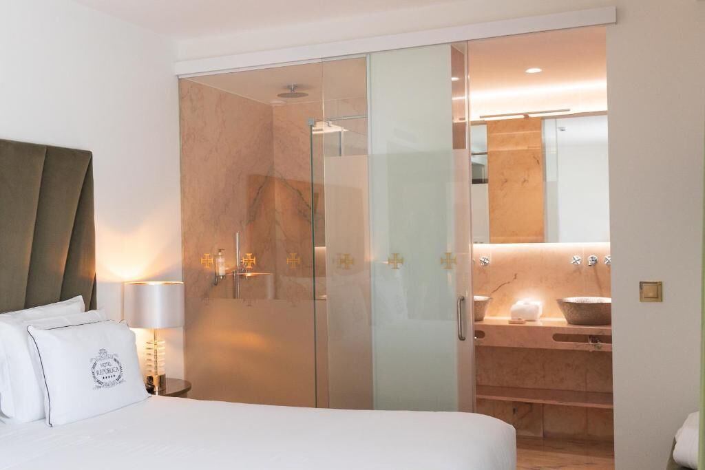 Quarto de hotel com cama, mesa de cabeceira, abajur, box de vidro no chuveiro e banheiro com pia grande e espelho.