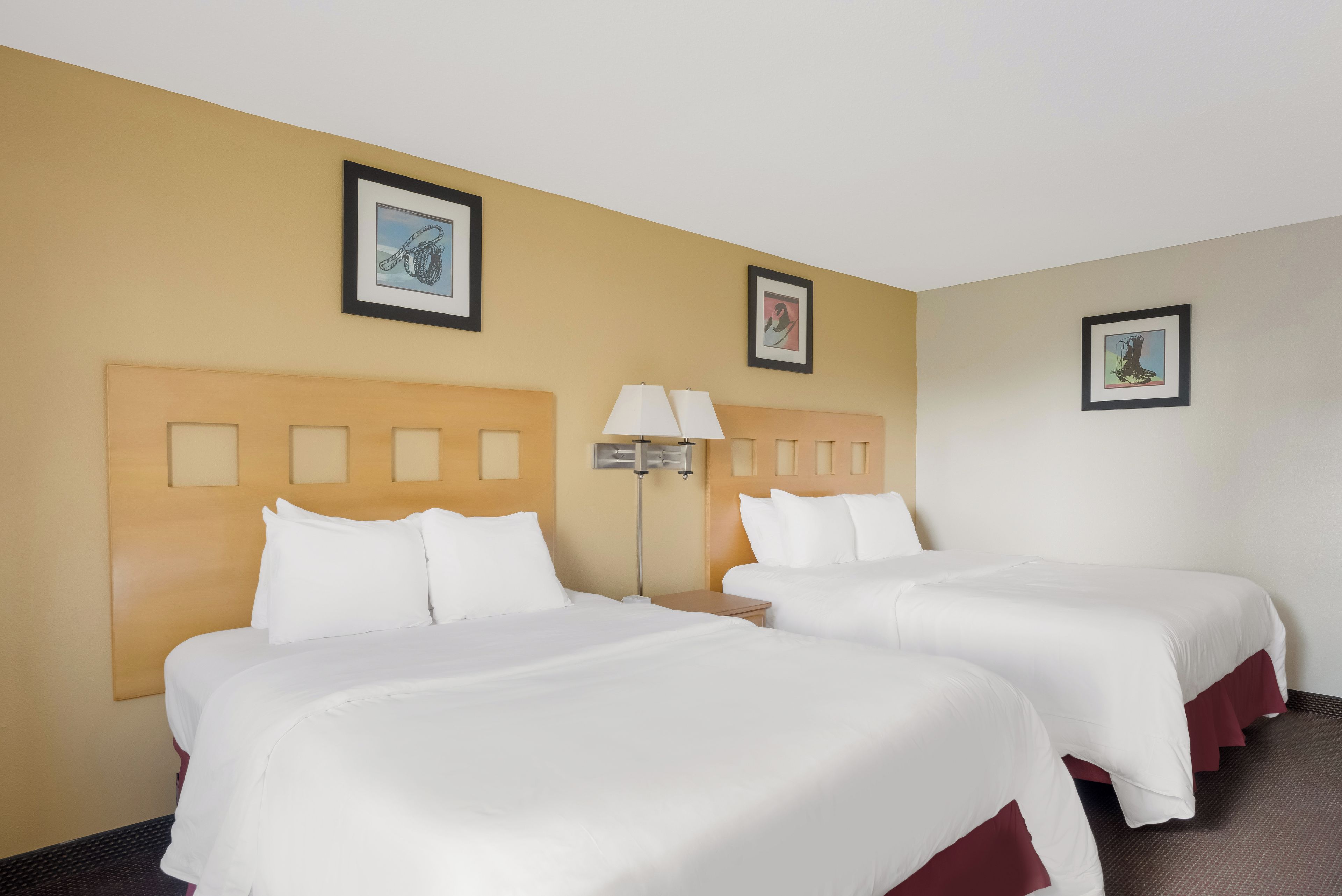 Quarto de hotel com duas camas, uma mesa de cabeceira, abajur e quadros nas paredes.