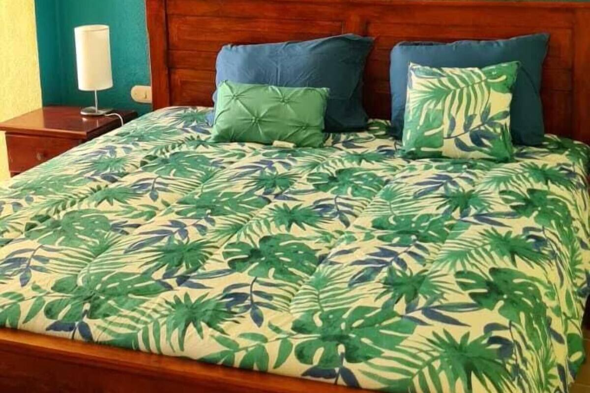 Quarto com cama de madeira, paredes verde-água, ventoinha no teto e decoração floral na parede.