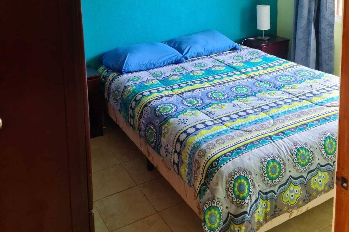 Quarto com parede verde-água, cama com colcha estampada colorida, duas gravuras de plantas emolduradas, um ventilador de teto e uma mesa de cabeceira com abajur.