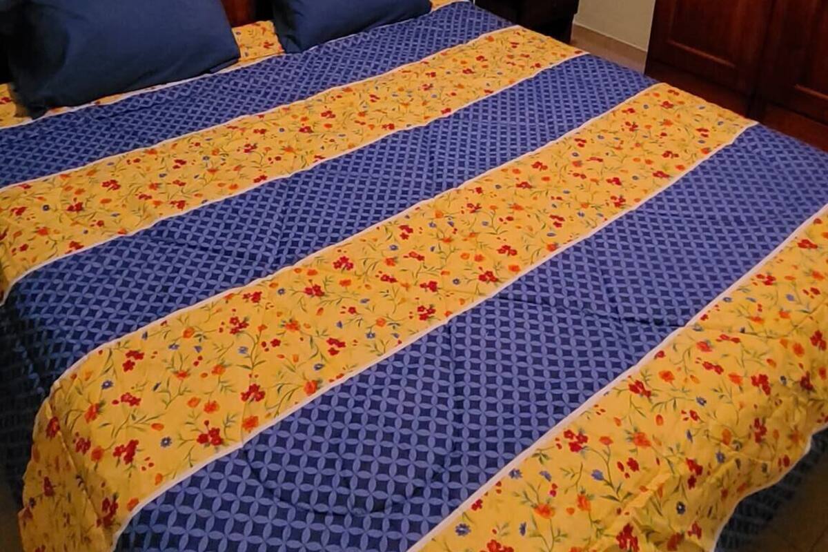 Quarto com parede azul, pintura floral, cama com colcha azul e amarela estampada, e mesa de cabeceira de madeira com abajur.