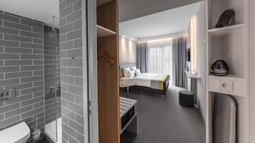 Quarto de hotel moderno com área de chuveiro revestida em azulejos cinzas, cama bem arrumada com mesa de cabeceira, pequeno espaço de estar e chaleira em uma prateleira.
