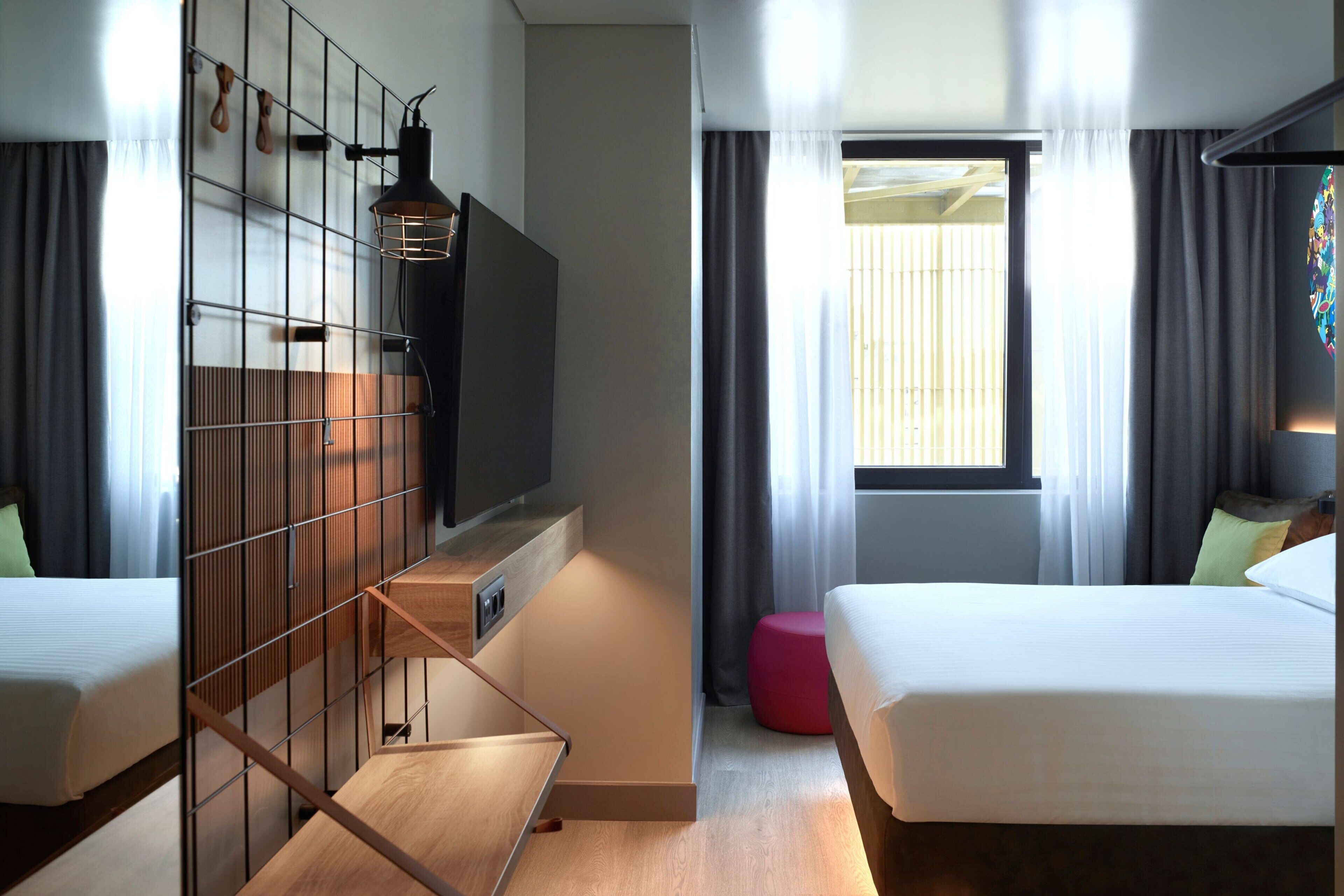 Quarto de hotel com duas camas, uma TV fixada em um painel de madeira, uma janela com cortinas e um banquinho pequeno.