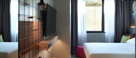Quarto de hotel com duas camas, uma TV fixada em um painel de madeira, uma janela com cortinas e um banquinho pequeno.