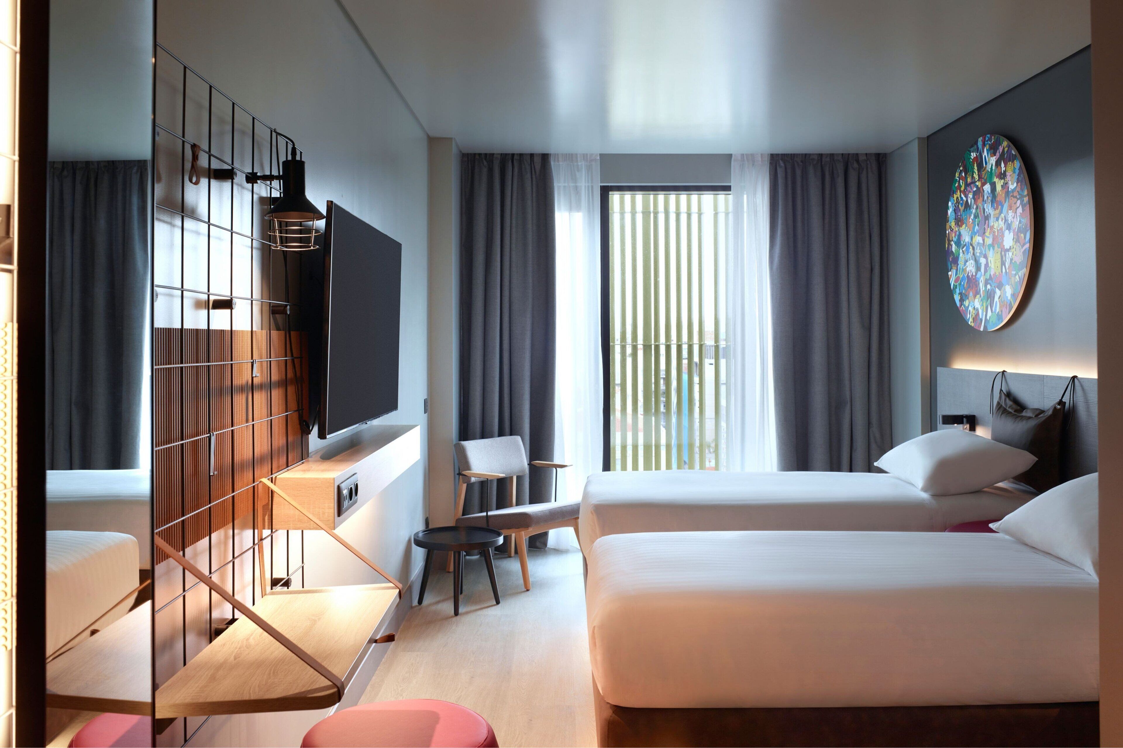 Quarto de hotel moderno com duas camas, televisão, escrivaninha e uma janela grande com cortinas.