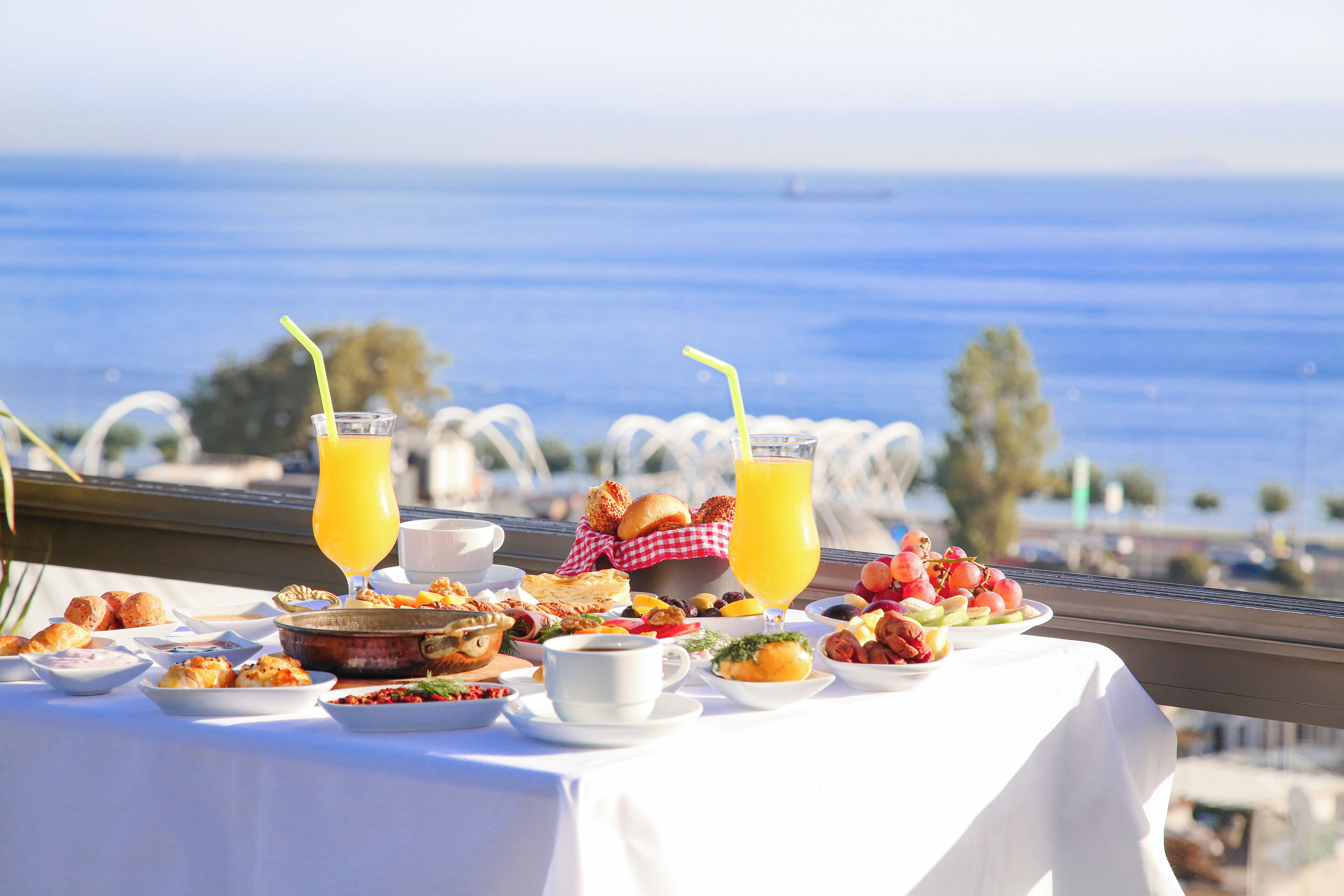 Mesa posta com itens de café da manhã e copos de suco de laranja, com vista panorâmica para o mar.
