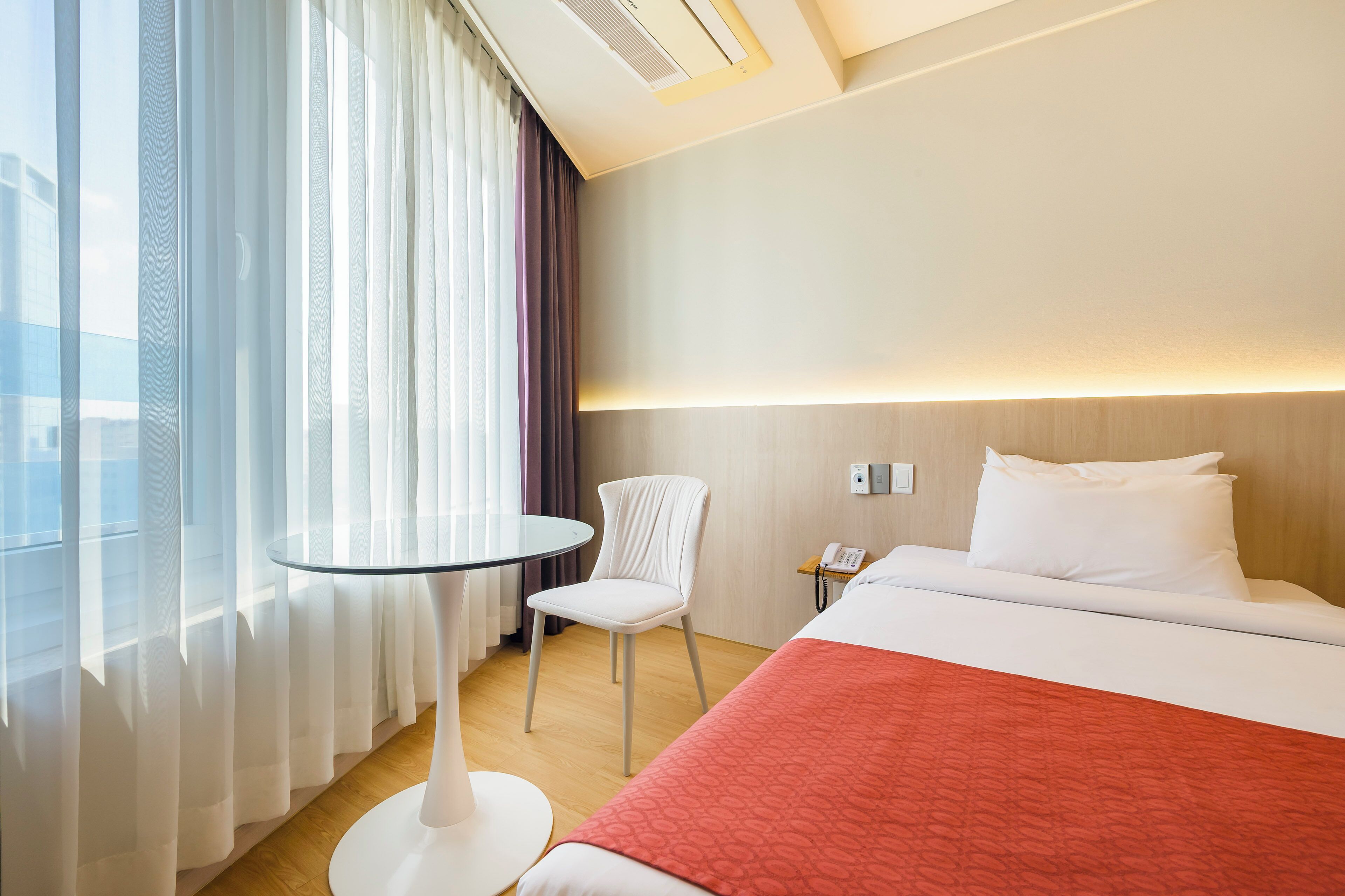 Quarto de hotel com cama, uma mesa pequena, uma cadeira e janela com cortinas leves.