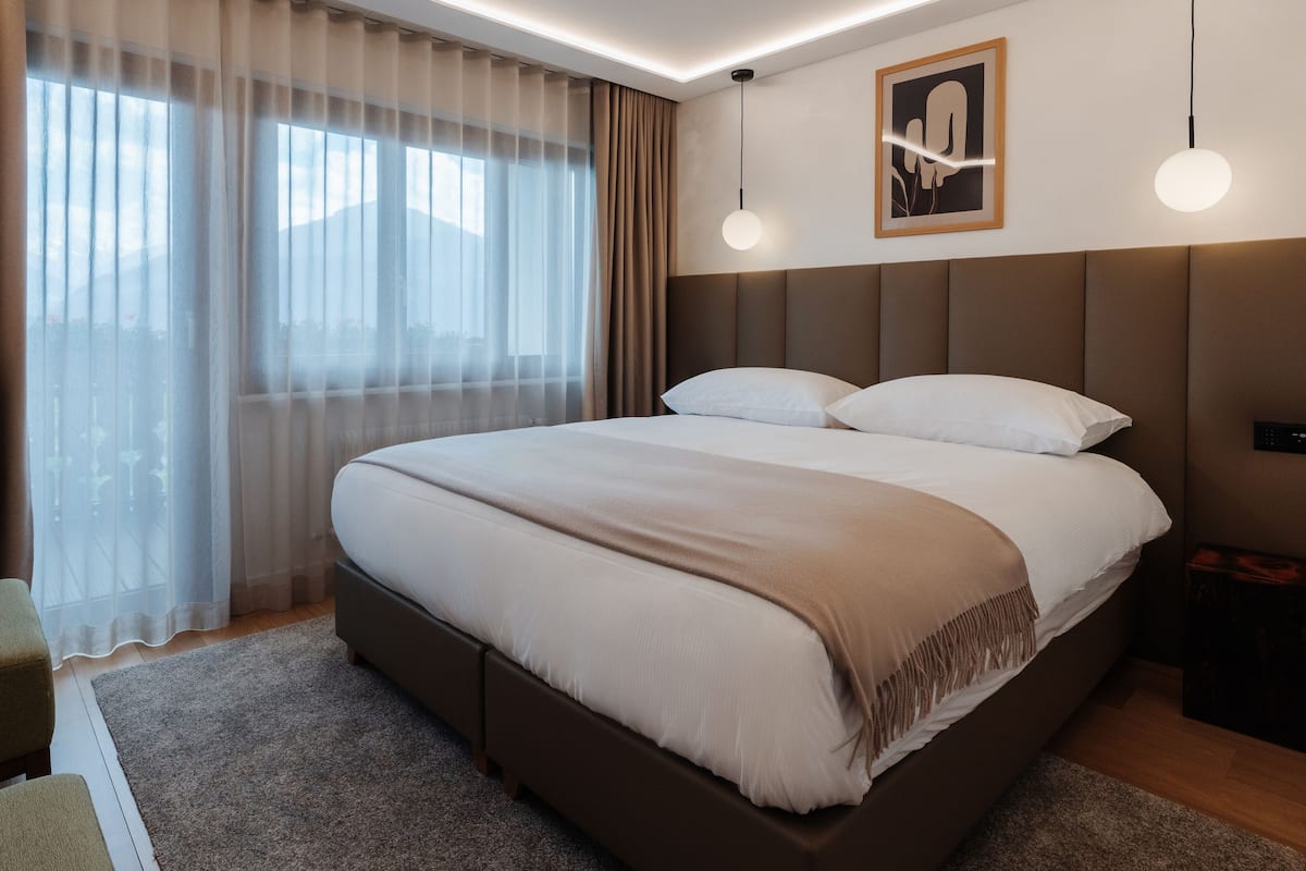 Quarto de hotel moderno com uma cama grande, vista para o exterior através de cortinas translúcidas e decoração minimalista.
