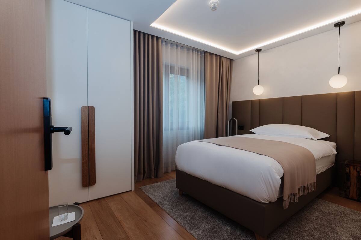 Quarto de hotel moderno com uma cama grande, mesa de cabeceira, guarda-roupa e uma janela com cortinas.